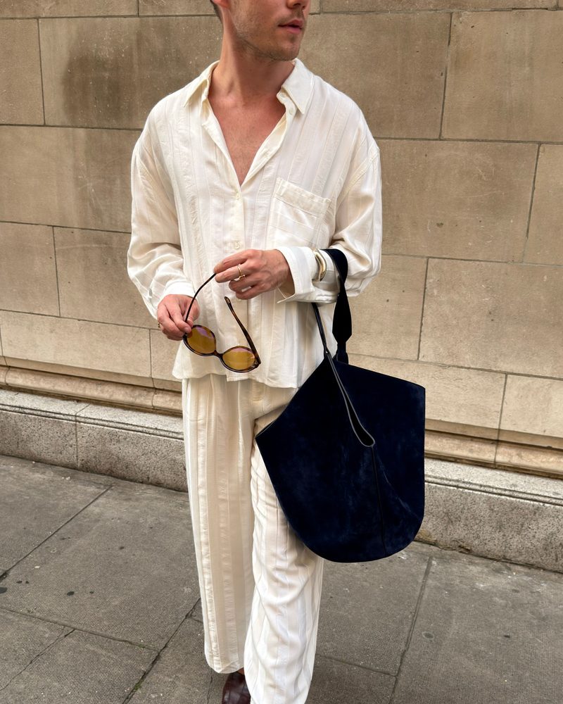 Man in a loose white linen outfit stands on a sidewalk. He holds sunglasses and a dark blue bag%2C exuding a casual%2C stylish vibe against a stone wall.