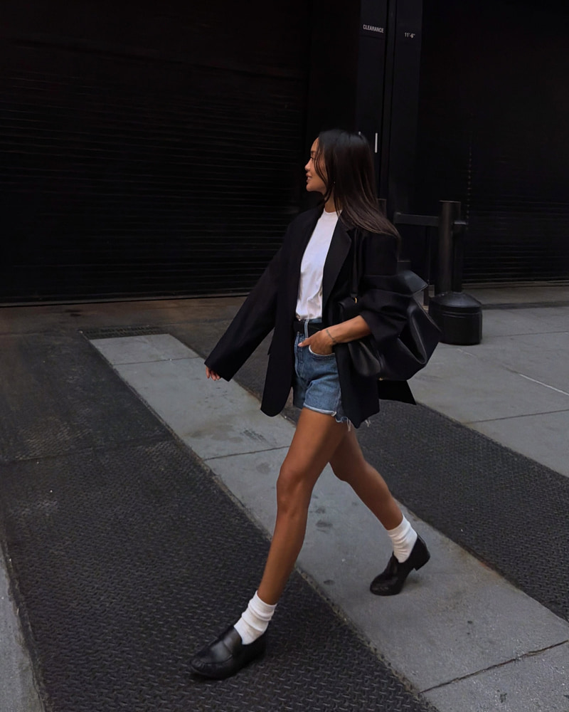 Woman walking in oversized black blazer%2C white t-shirt%2C denim shorts%2C white socks%2C and black leather loafers in urban setting
