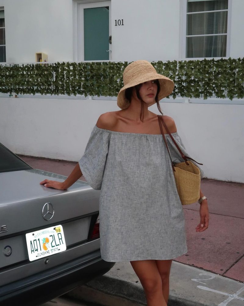A woman in a grey off-shoulder dress and straw hat stands casually by a parked car%2C carrying a straw bag. The setting is a quiet urban street.