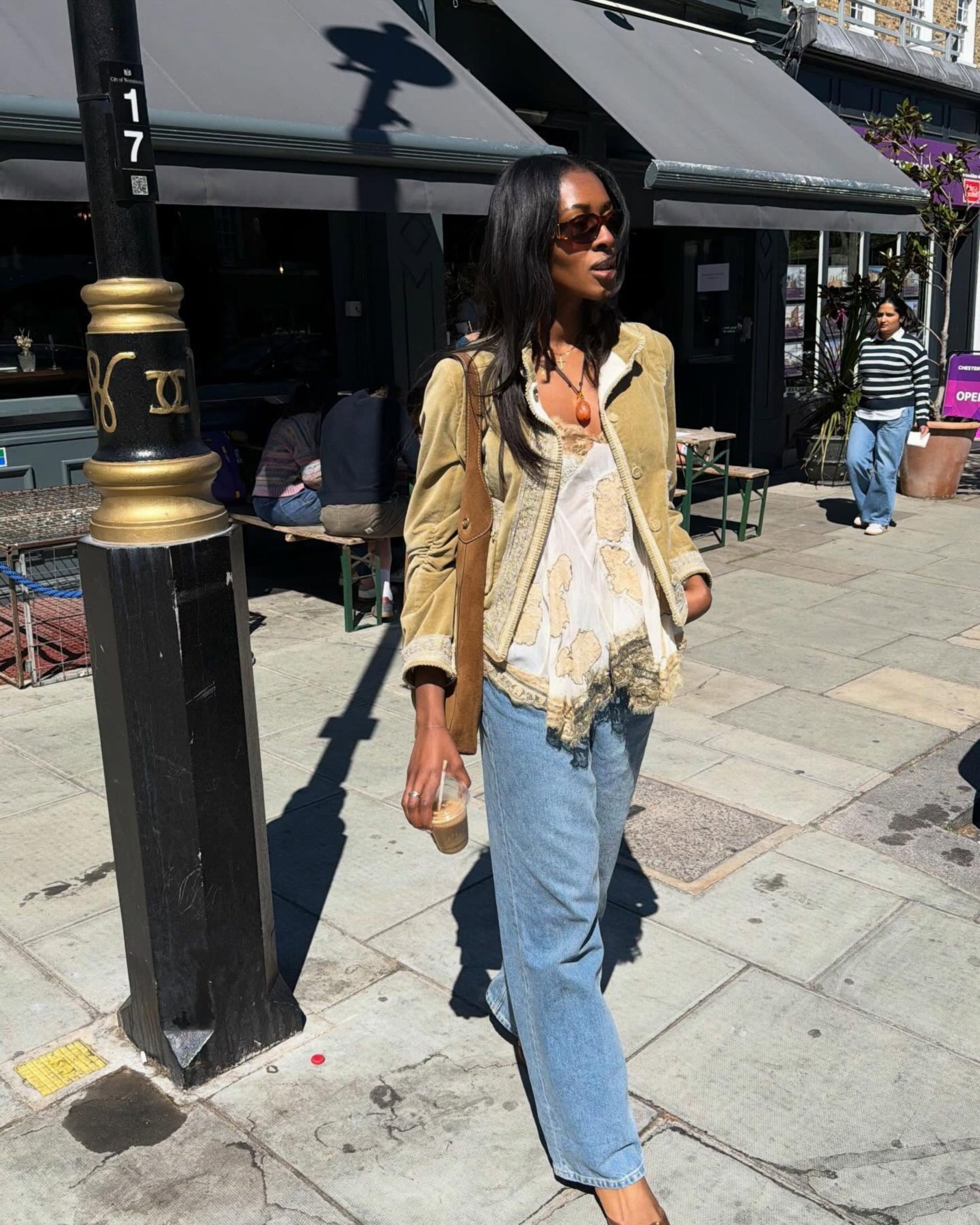 Woman wearing a beige shearling jacket over a cream lace-trimmed blouse paired with light blue wide-leg jeans and brown sunglasses walking on a sunny city street