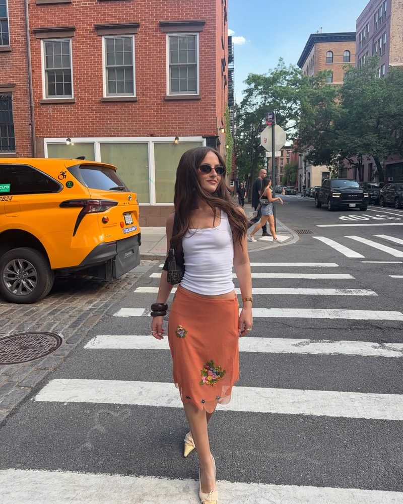 A woman confidently walks across a city street crosswalk. She wears sunglasses%2C a white tank top%2C and an orange floral skirt. A yellow taxi and red brick buildings line the background%2C creating an urban scene.