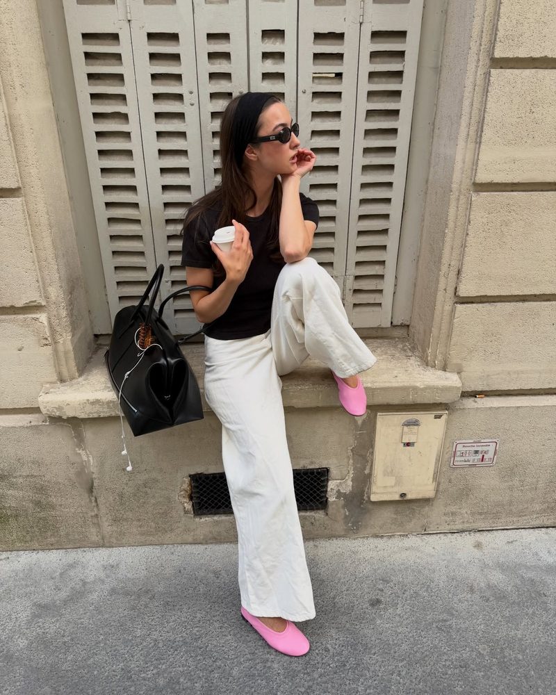 Woman in casual outfit leans against a cream wall%2C wearing sunglasses and pink shoes. She holds a coffee cup and has a large black bag beside her.