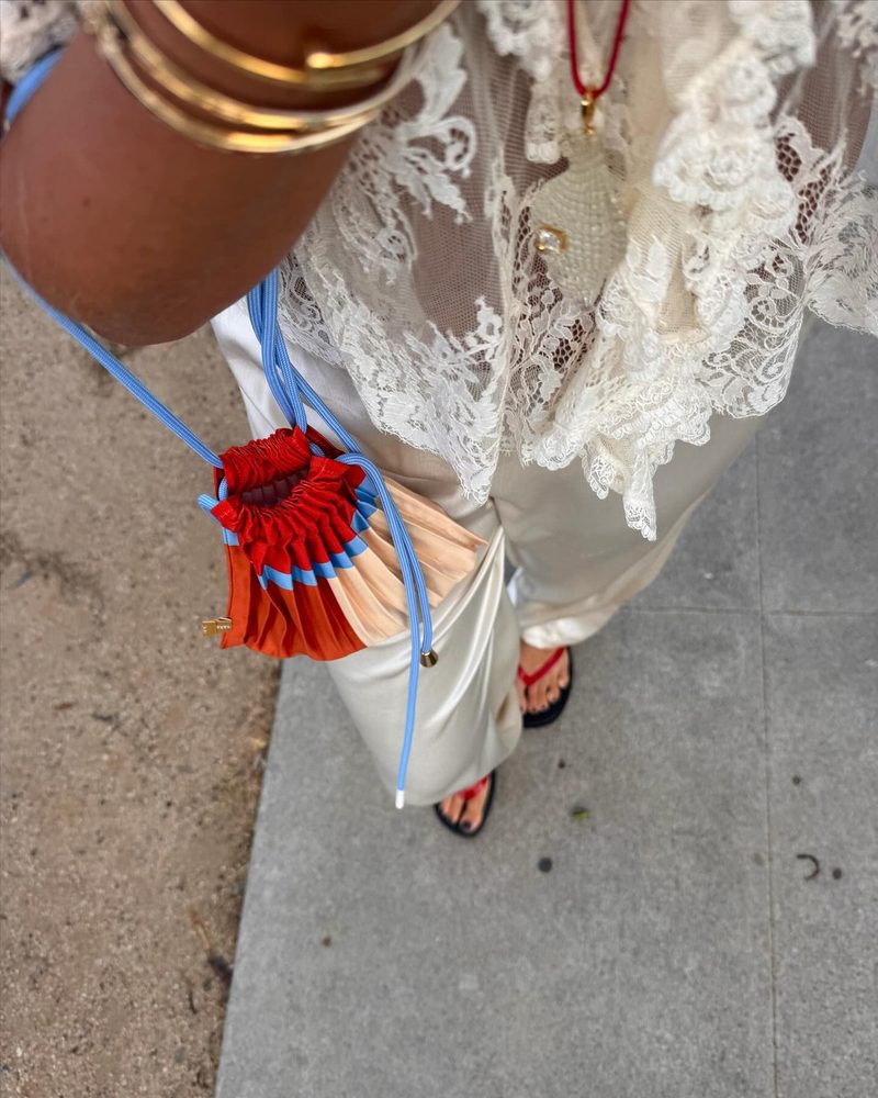 Overhead view of a person wearing a lace top%2C white silk pants%2C and black sandals%2C holding a colorful drawstring bag. Accessories include gold bracelets.