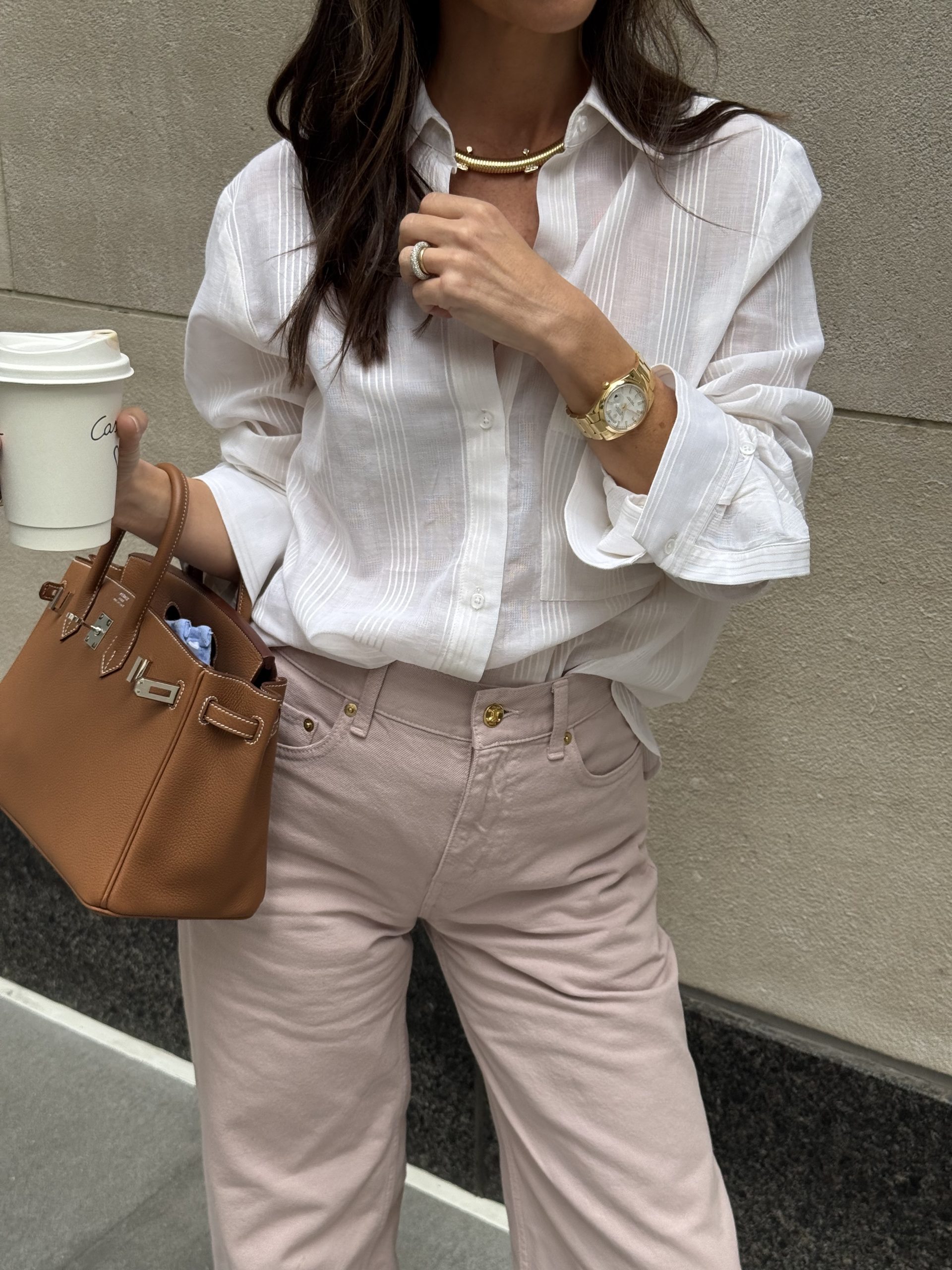 Woman in a white blouse and light pink pants holds a brown handbag and coffee cup. Wears a gold watch and necklace%2C creating a chic%2C sophisticated look.