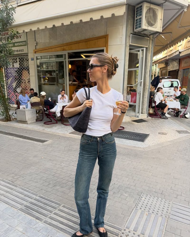 Woman in casual attire holds a drink and handbag on a cobblestone street. Nearby%2C people dine outside a caf%C3%A9%2C creating a lively%2C relaxed ambiance.