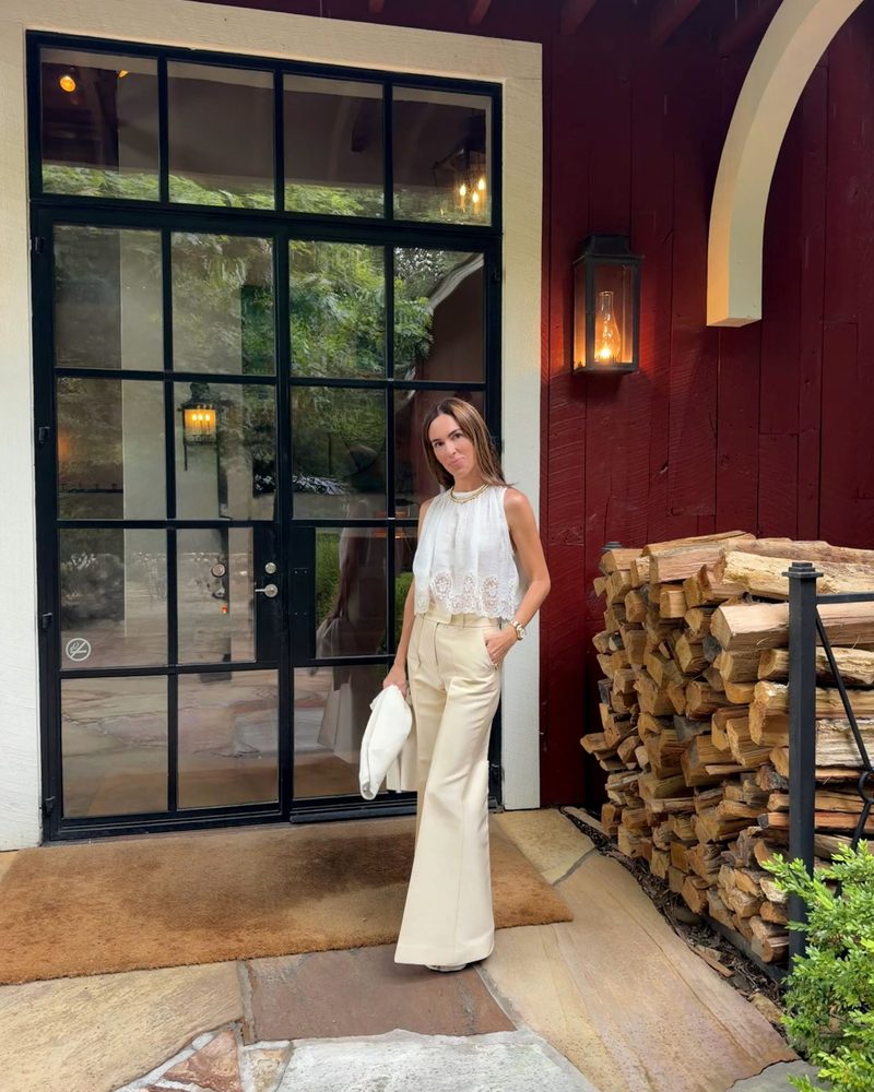Woman in a white lace top and cream pants stands by a pile of firewood next to a glass door. The setting feels rustic and serene.
