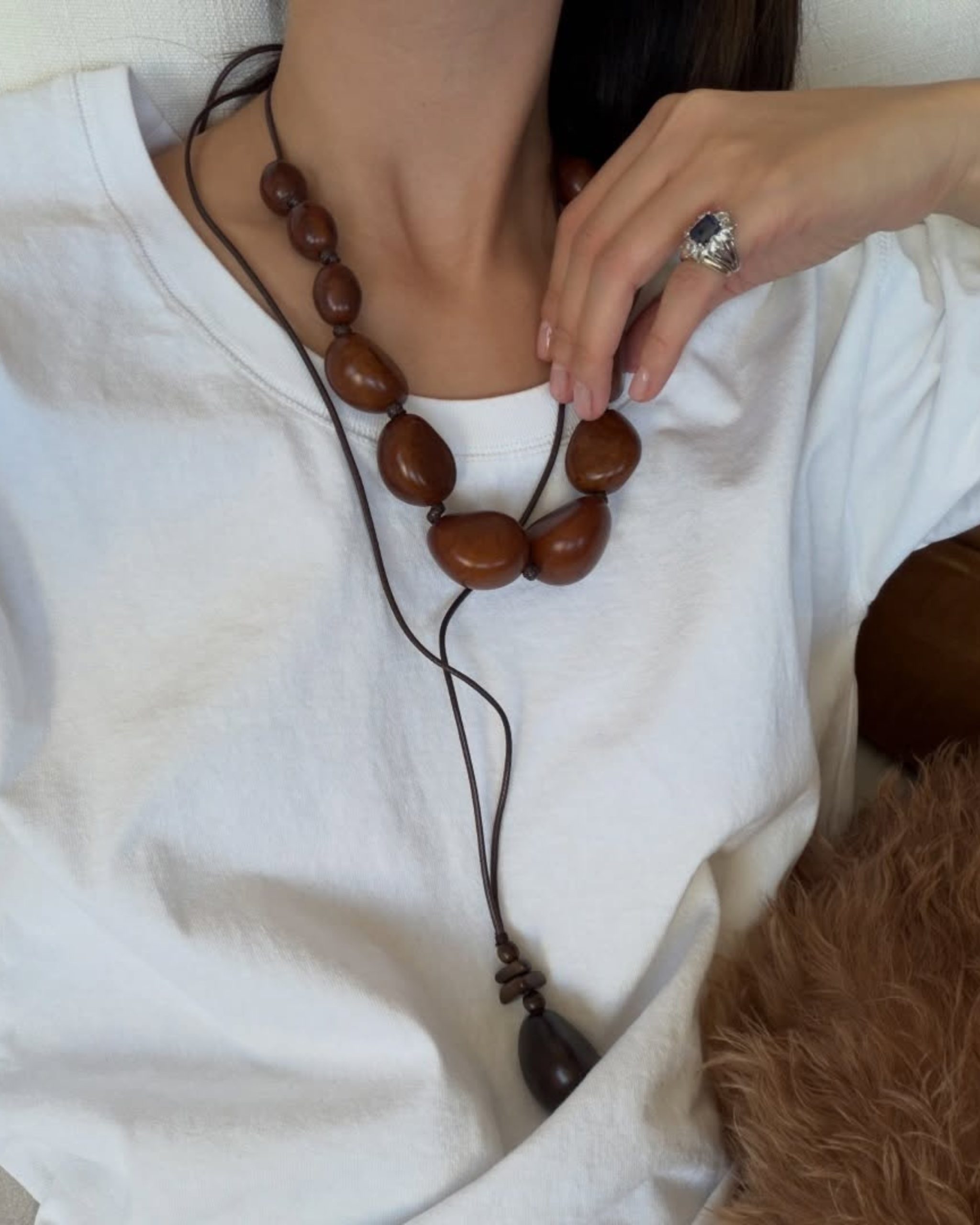 A person wearing a white t-shirt is lounging comfortably. They have a chunky%2C beaded wooden necklace. Their hand rests near their collar%2C displaying a large ring. A cozy brown cushion is partially visible%2C adding warmth to the relaxed setting.