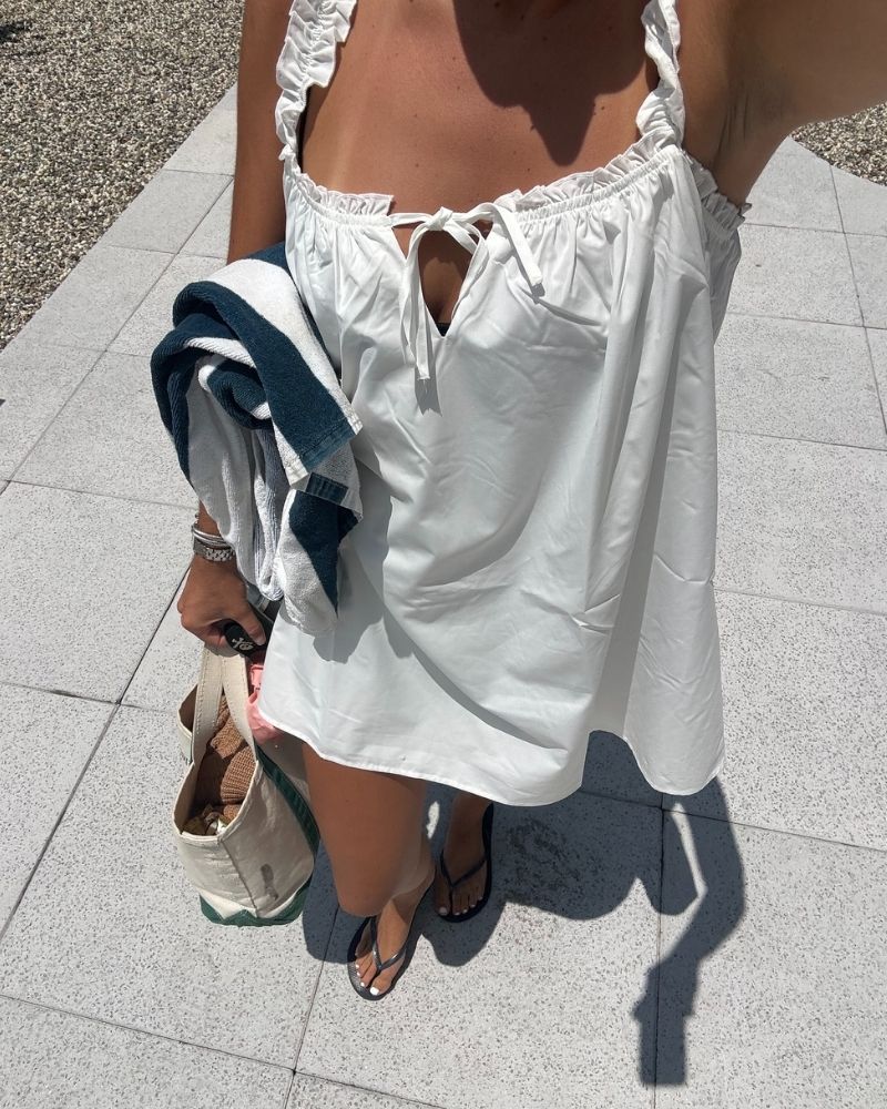 White babydoll dress with ruffled straps and front tie detail styled with black flip-flops and a tote bag for summer beachwear