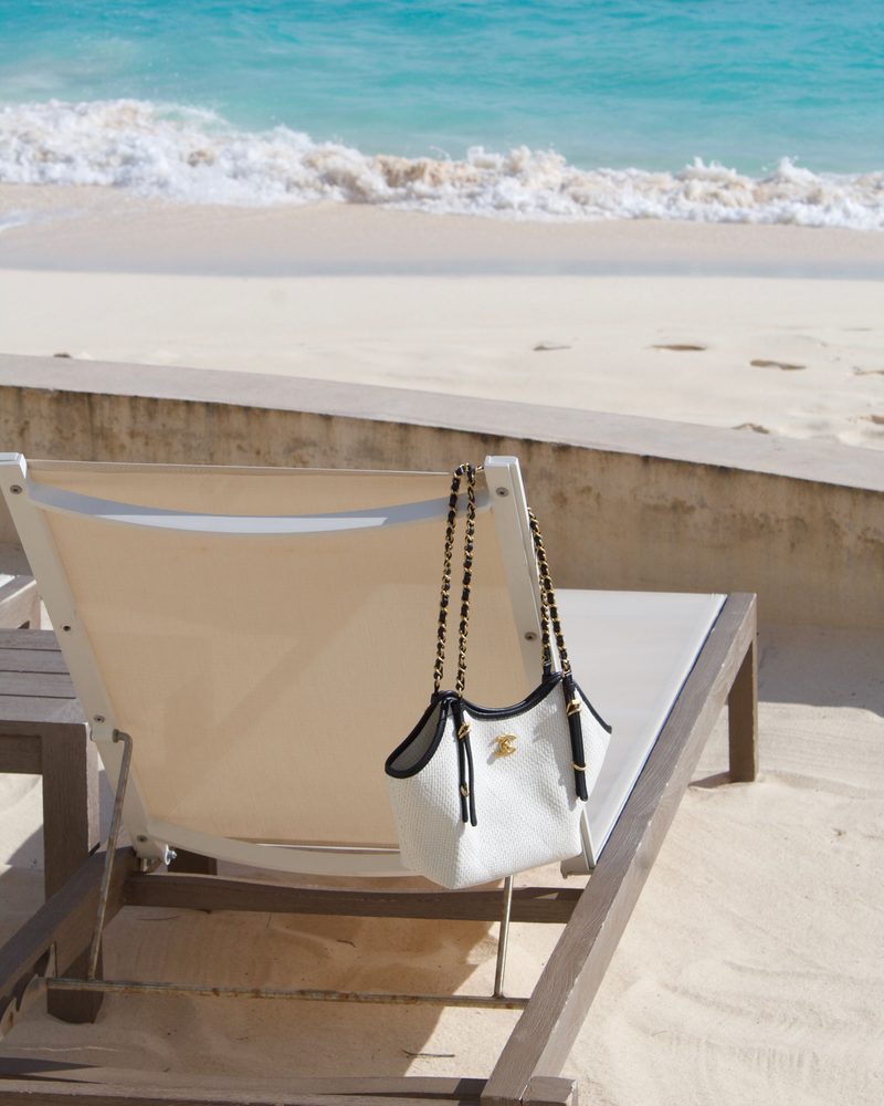 White leather designer handbag with black trim and gold chain straps hanging on a beige beach lounge chair by turquoise ocean waves