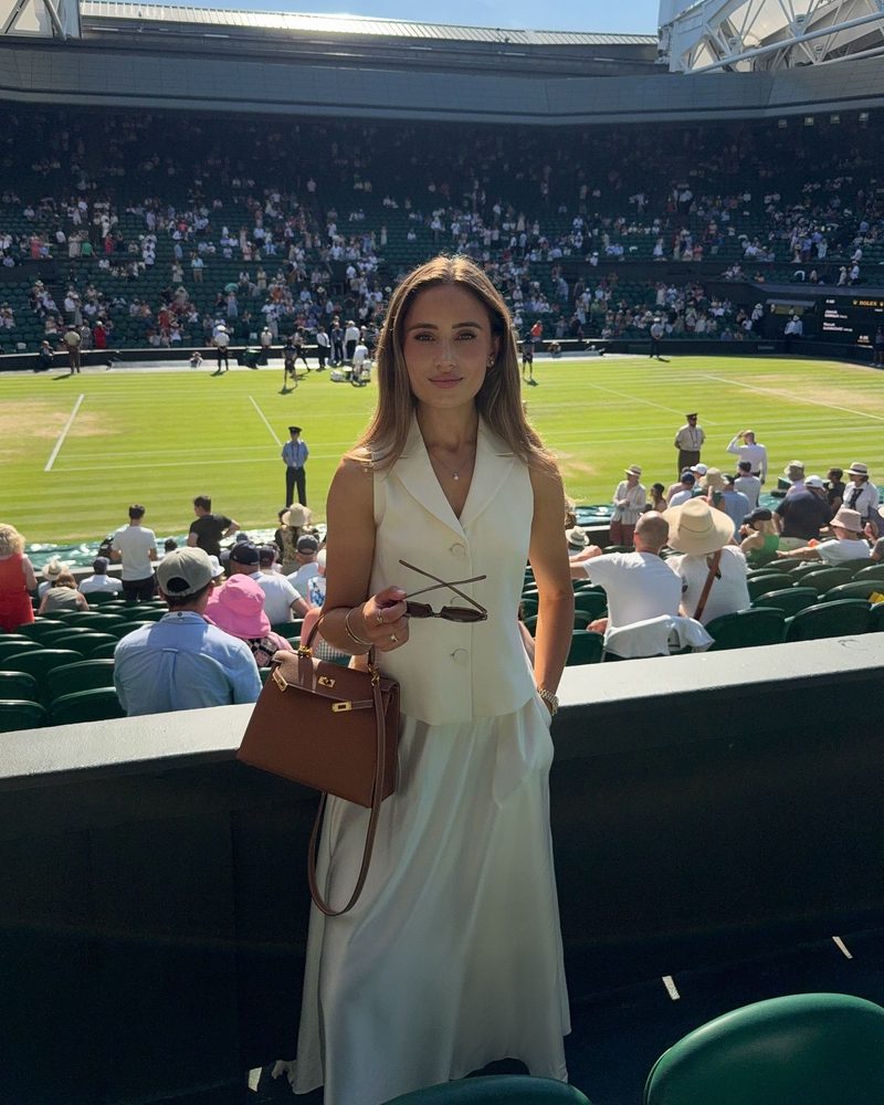 Elegant outfit at a tennis match features a chic white vest dress paired with a stylish brown handbag and sunglasses%2C set against a vibrant crowd background