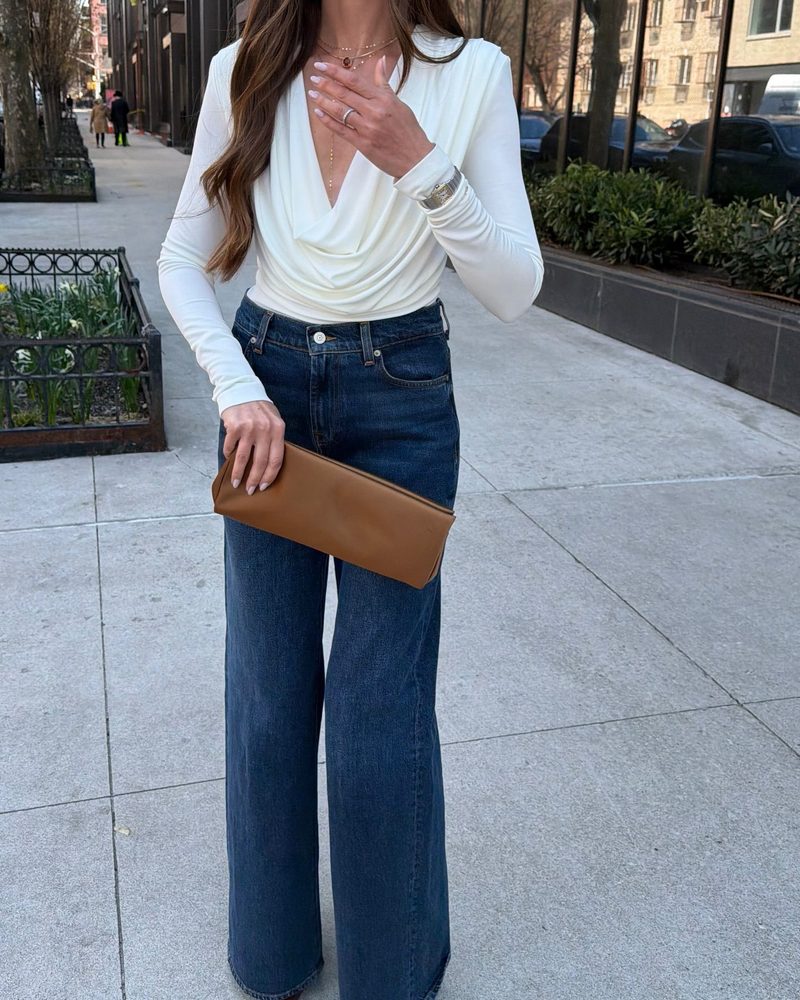 Elegant draped white blouse paired with high-waisted dark denim flares%2C complemented by a chic brown clutch and a layered necklace