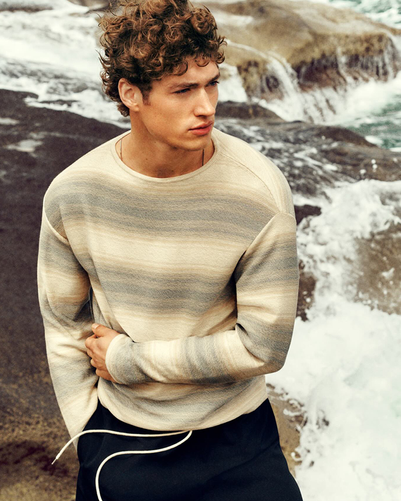 Man in a striped knit sweater and relaxed black drawstring pants on a rugged rocky shoreline in coastal scenery