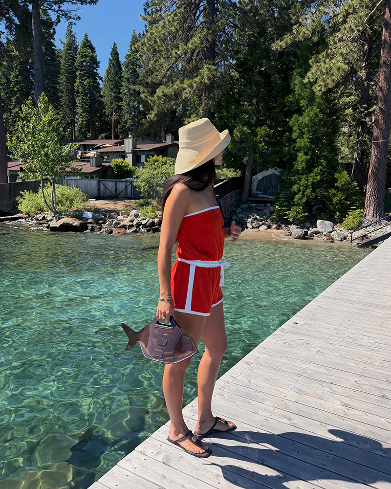 Red terry cloth romper with a wide-brim straw hat%2C fish-shaped handbag and flip-flops%2C styled on a lakeside dock