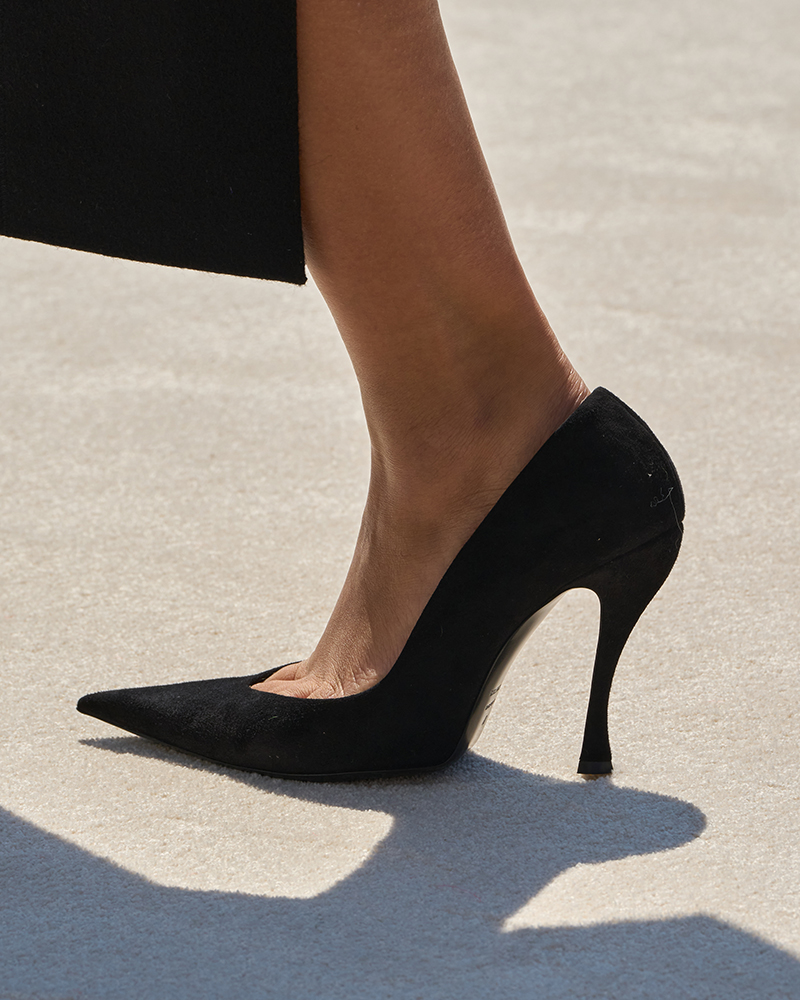 Close-up of black suede pointed-toe pumps with sculptural stiletto heel%2C styled on a sunlit stone surface