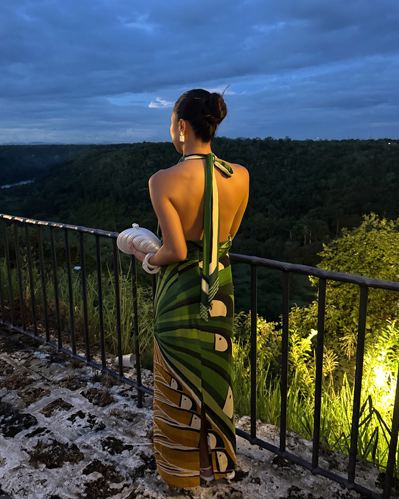 Green printed halter maxi dress with an open-back design and white clutch%2C overlooking a lush valley at dusk