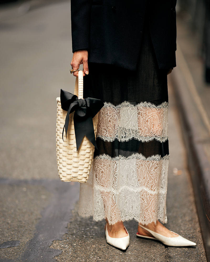 Close-up of a black lace maxi skirt with white lace panels%2C woven basket bag and white slingback flats on a city street