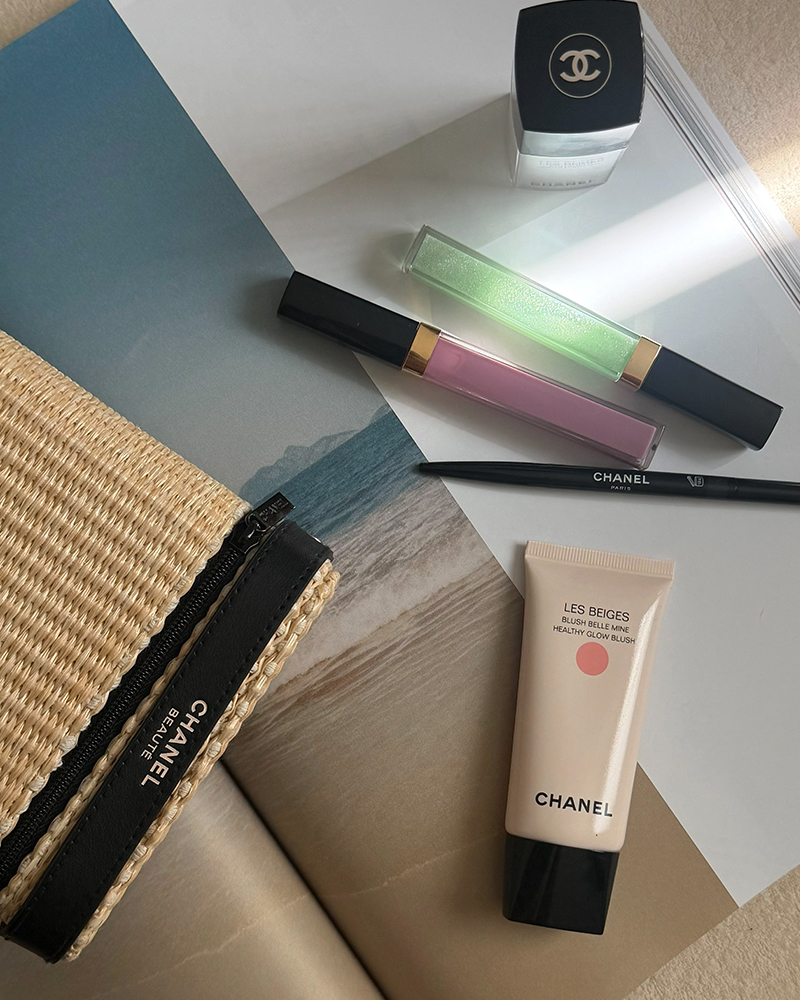 Chanel beauty essentials featuring blush%2C lip gloss%2C eyeliner and woven clutch%2C styled on magazines in a flat lay