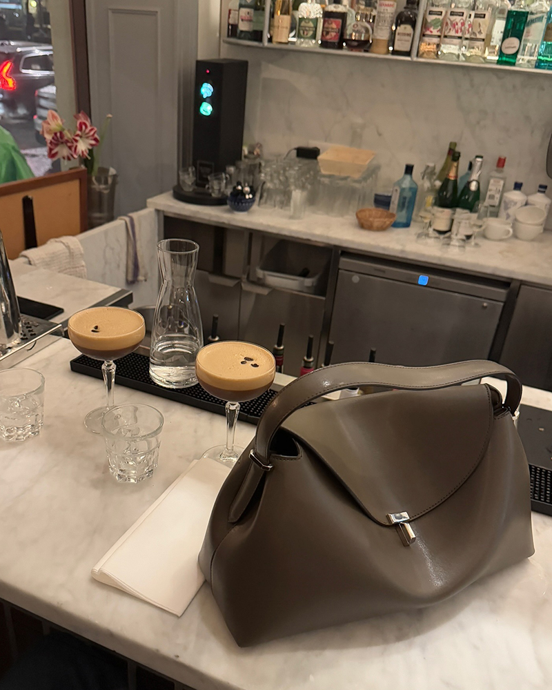 Taupe leather top-handle handbag with sculptural flap closure on a marble bar counter beside espresso martinis
