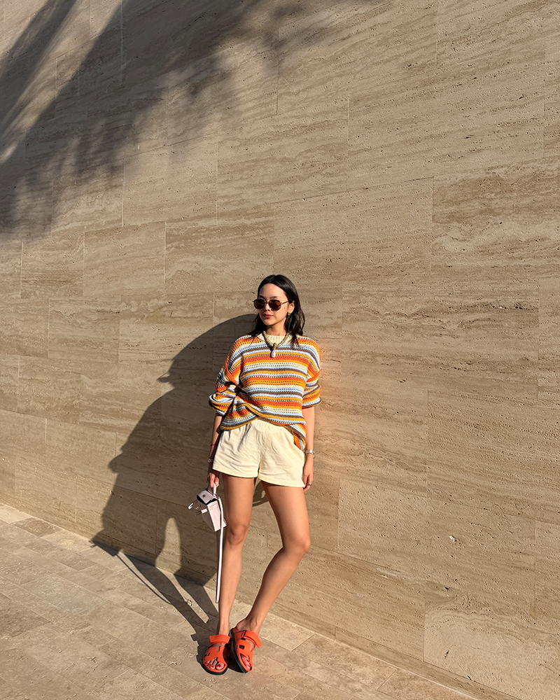 Striped crochet sweater paired with cream shorts%2C orange sandals and sunglasses%2C styled against a sunlit stone wall for summer