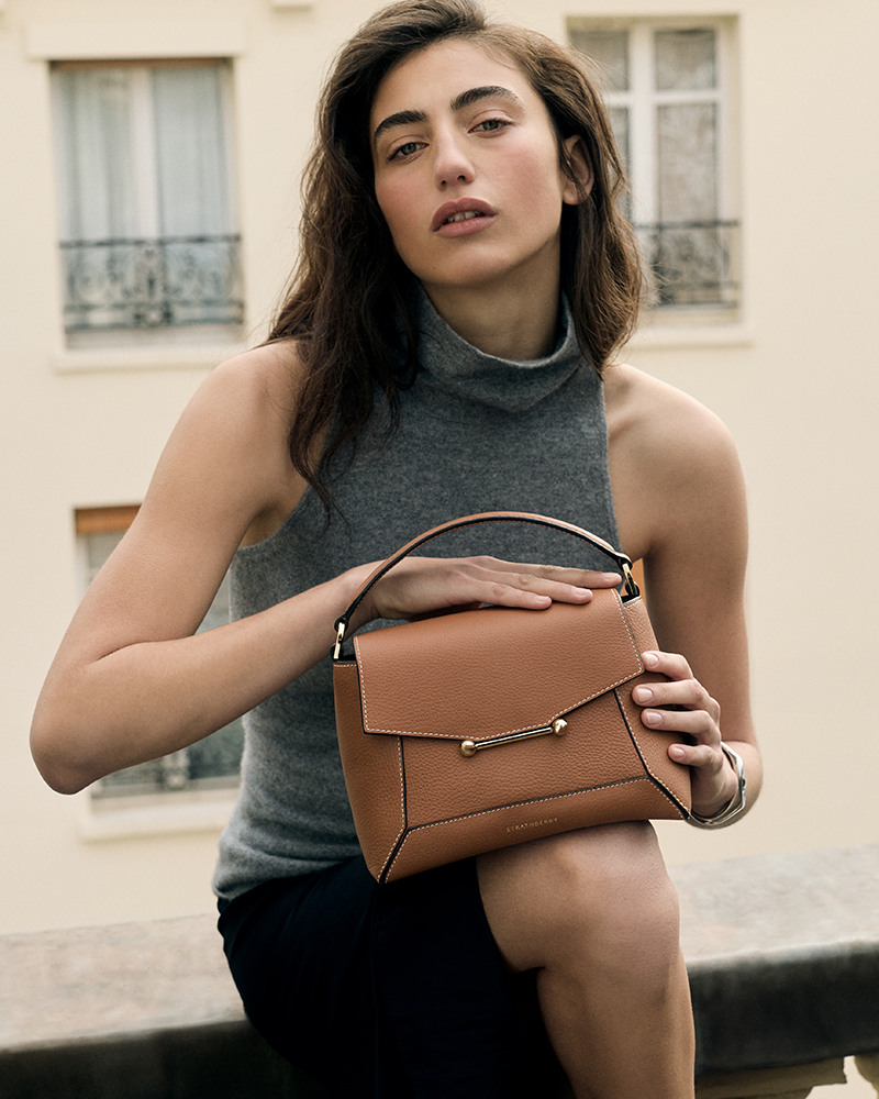 Strathberry tan leather top-handle handbag paired with a gray sleeveless knit top%2C photographed on an urban terrace backdrop