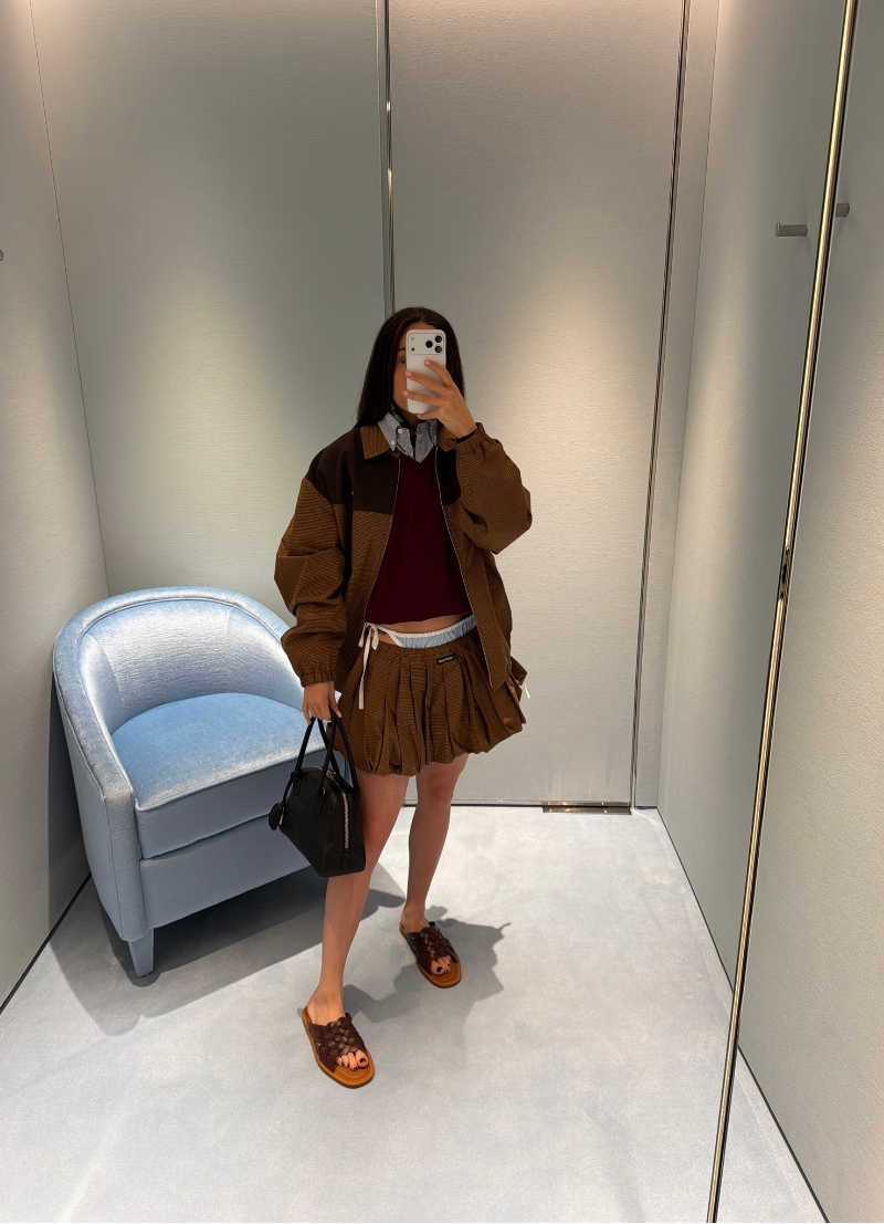 Miu Miu mirror selfie featuring a brown bomber jacket, pleated mini skirt, knit top and woven leather slides in a fitting room