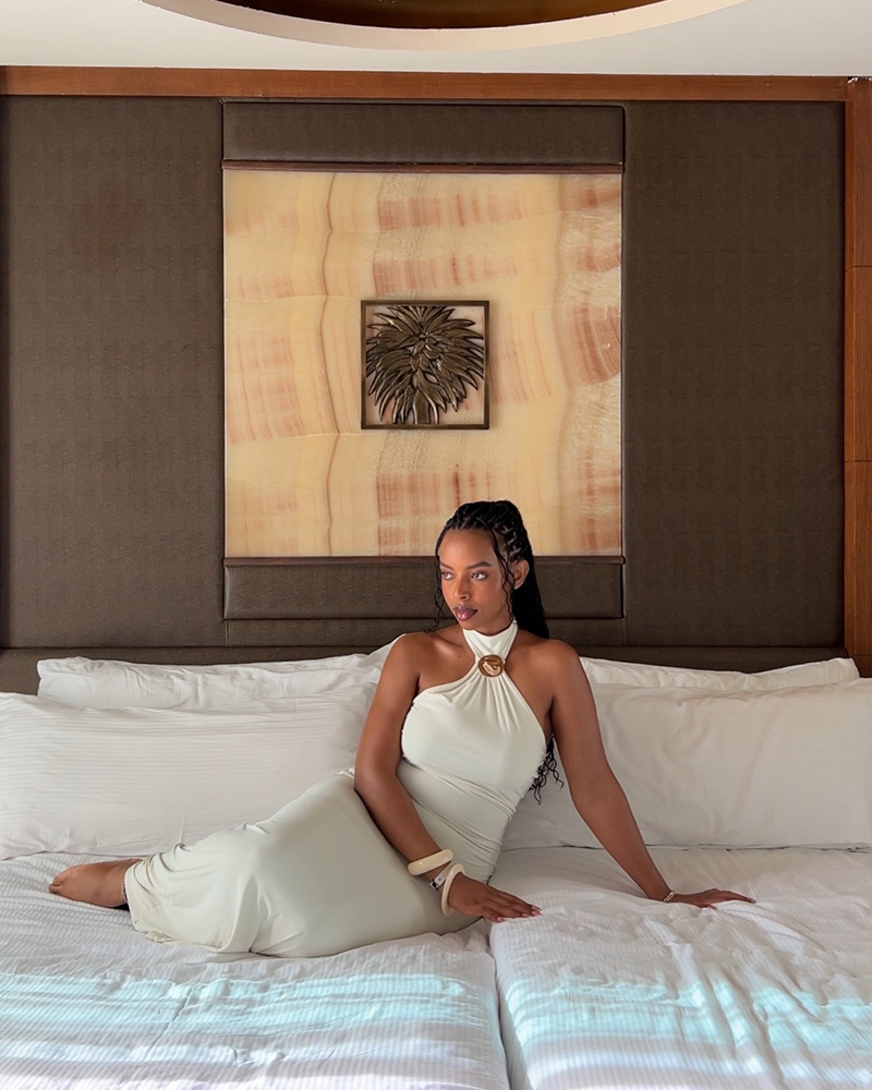 Cream halter maxi dress with gold statement neckline and bangles%2C styled on a hotel bed in a luxe resort setting