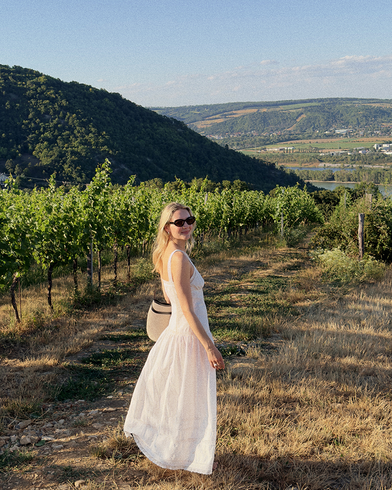 White cotton summer maxi dress styled with black sunglasses and a woven shoulder bag in a scenic vineyard landscape
