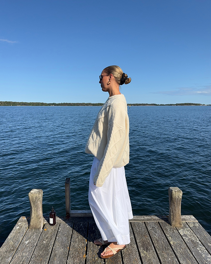 Cream cable-knit sweater paired with a flowing white maxi skirt and sandals%2C standing on a lakeside dock under a clear blue sky