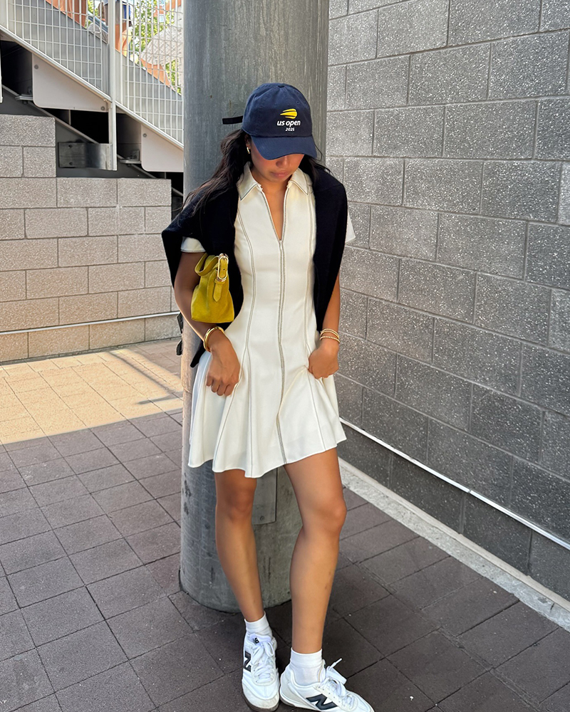 Model in a white tennis dress%2C US Open cap and sneakers with a yellow shoulder bag at the stadium