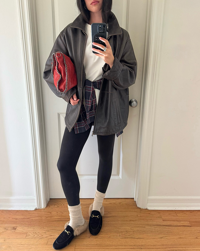 Mirror selfie featuring an oversized leather jacket%2C black leggings%2C loafers and a red chain-trim clutch in a minimalist hallway%0A