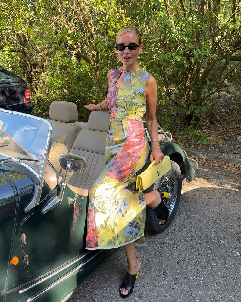 Printed sleeveless maxi dress with a colorful floral pattern%2C yellow clutch and sunglasses%2C styled beside a vintage convertible outdoors