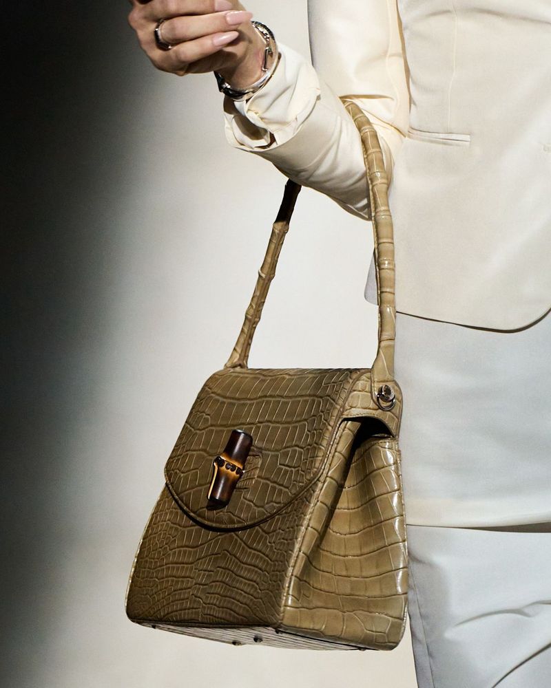 Gucci beige crocodile leather handbag with bamboo clasp and structured top handle held by model in cream suit