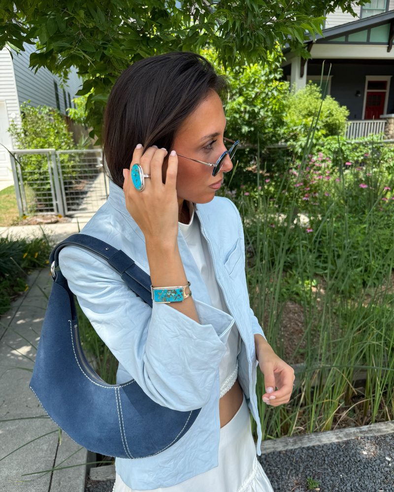 Light blue cropped jacket paired with a white crop top and tiered skirt%2C accessorized with a navy suede handbag and turquoise statement jewelry.