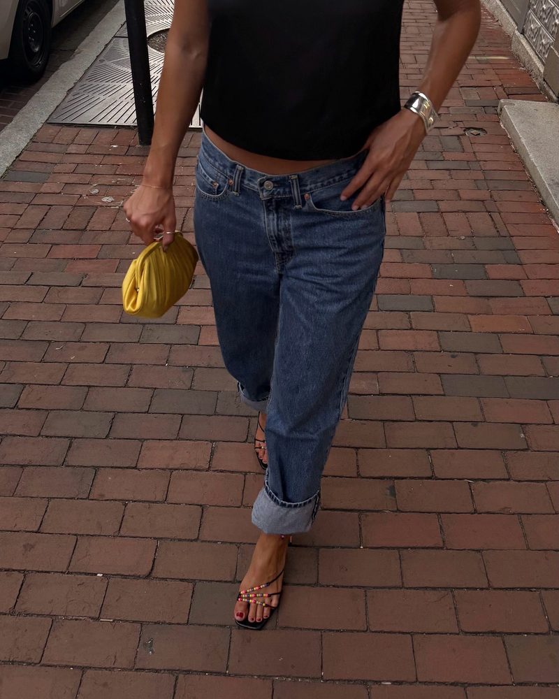 Casual outfit featuring rolled-up blue denim jeans%2C black cropped top%2C colorful beaded strappy sandals%2C silver cuff bracelet%2C and a small yellow clutch bag