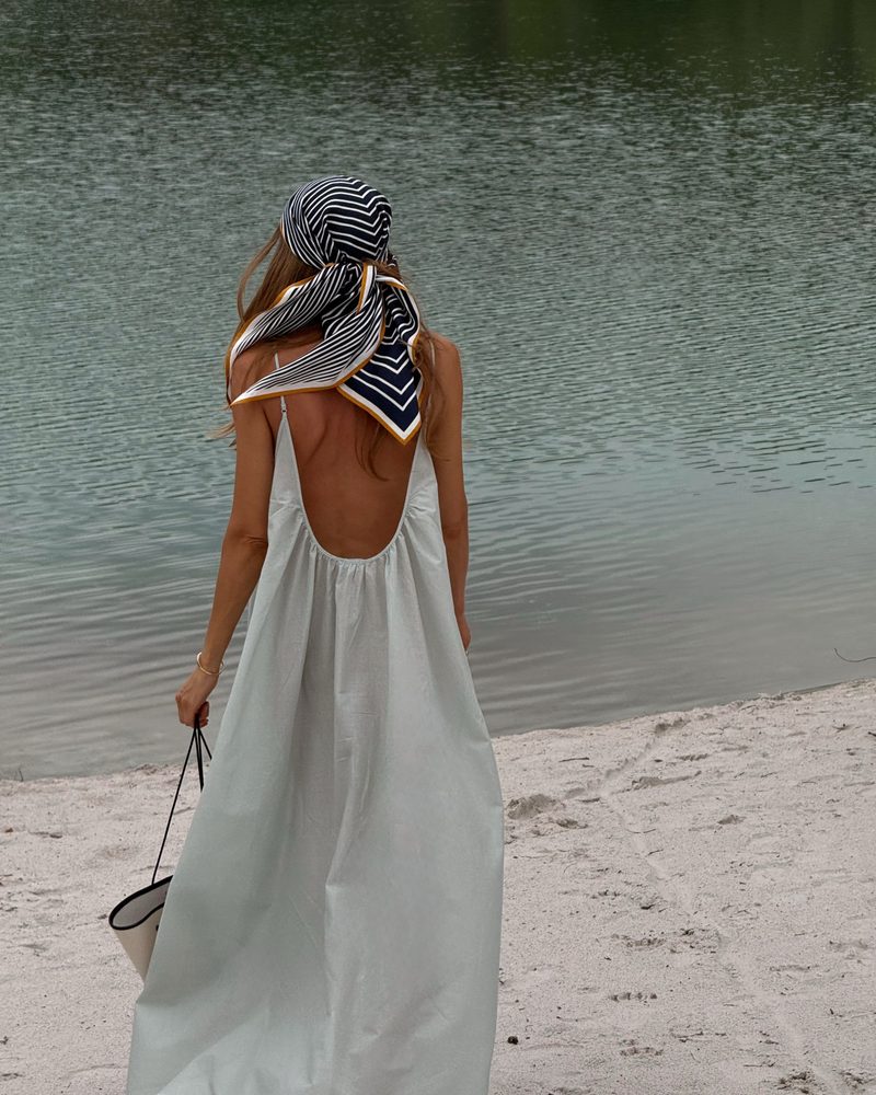 Woman wearing a white backless maxi dress with thin straps and a navy striped silk headscarf standing by a lakeside beach