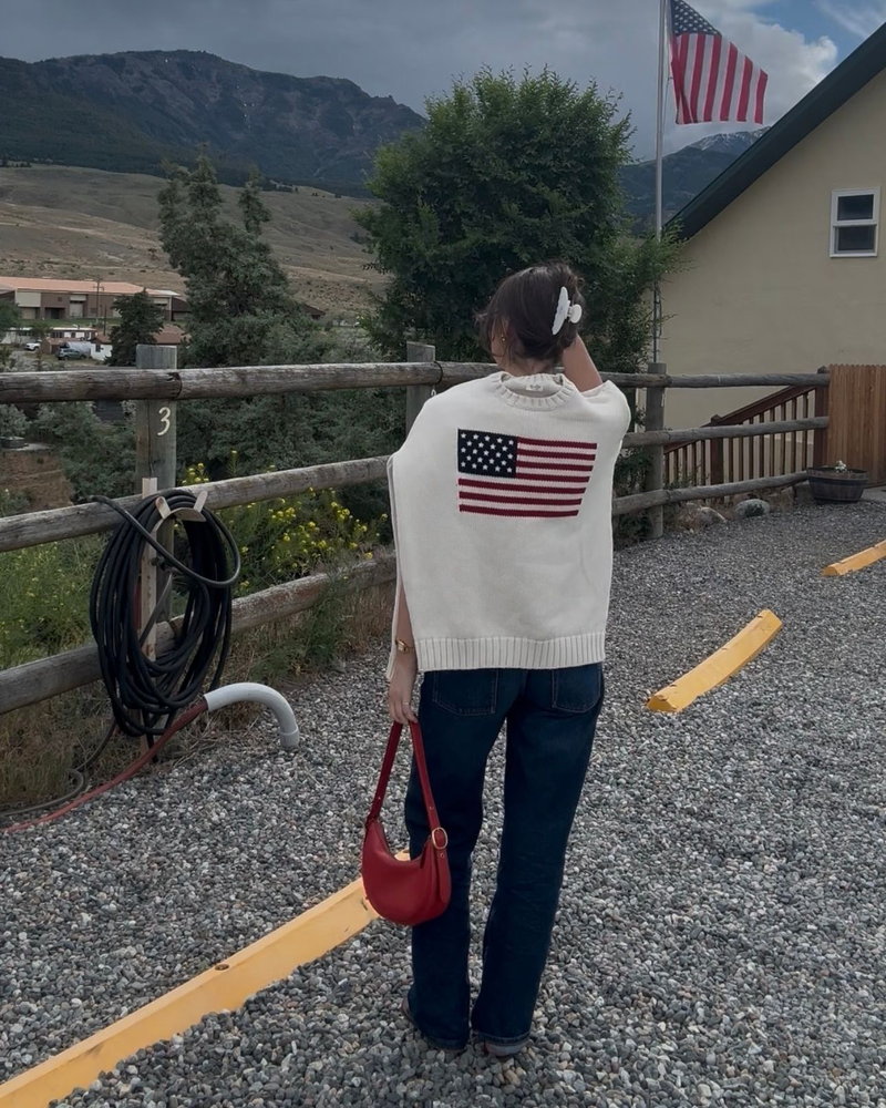 Woman wearing a white knit sweater with an American flag design%2C dark jeans%2C and a red handbag in a rustic outdoor setting%0A%0A