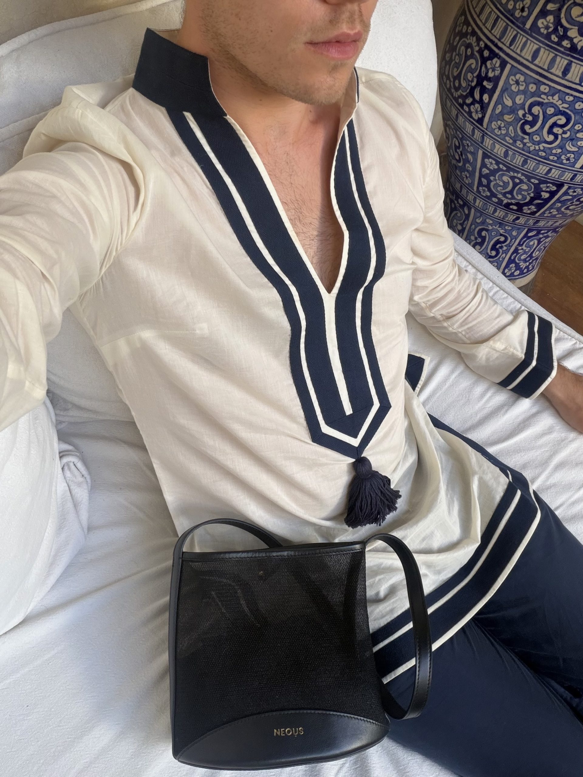 A person wearing a beige shirt with navy accents and tassel lounges on a white couch. A black bag rests nearby. Decorative blue vase in the background.