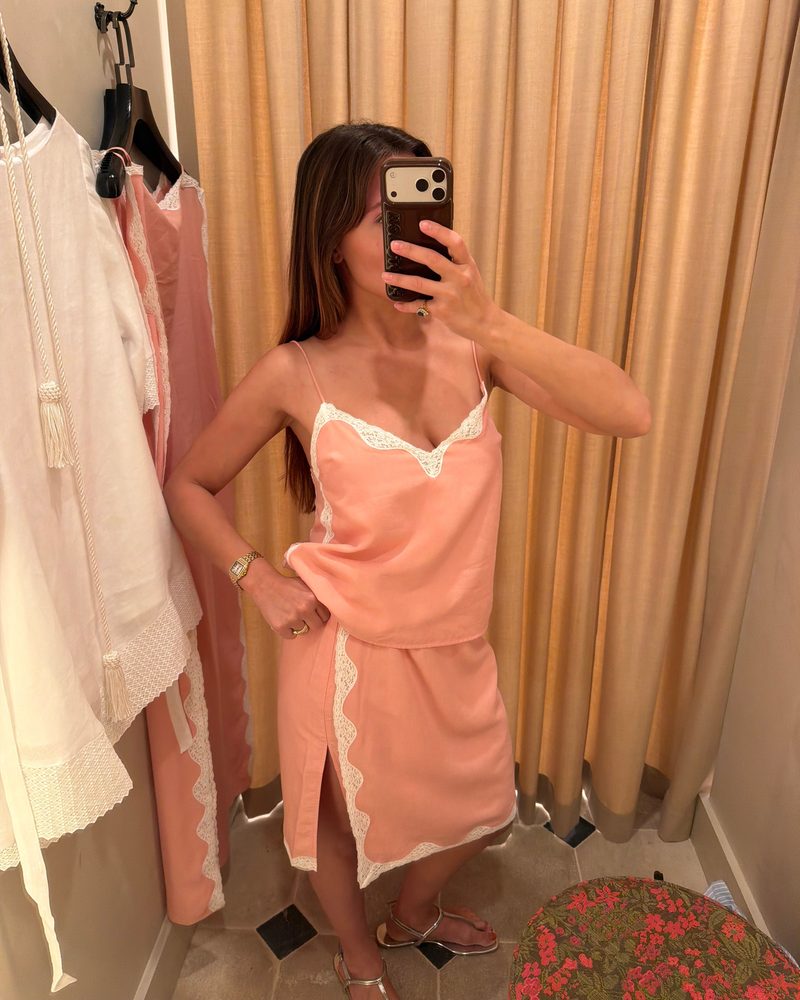 Woman wearing a peach satin camisole and skirt set with white lace trim%2C styled with gold sandals and a gold watch in a boutique fitting room