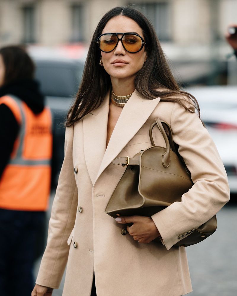 Woman wearing a beige double-breasted blazer with gold buttons%2C oversized amber sunglasses%2C gold choker necklace%2C and carrying a taupe leather handbag