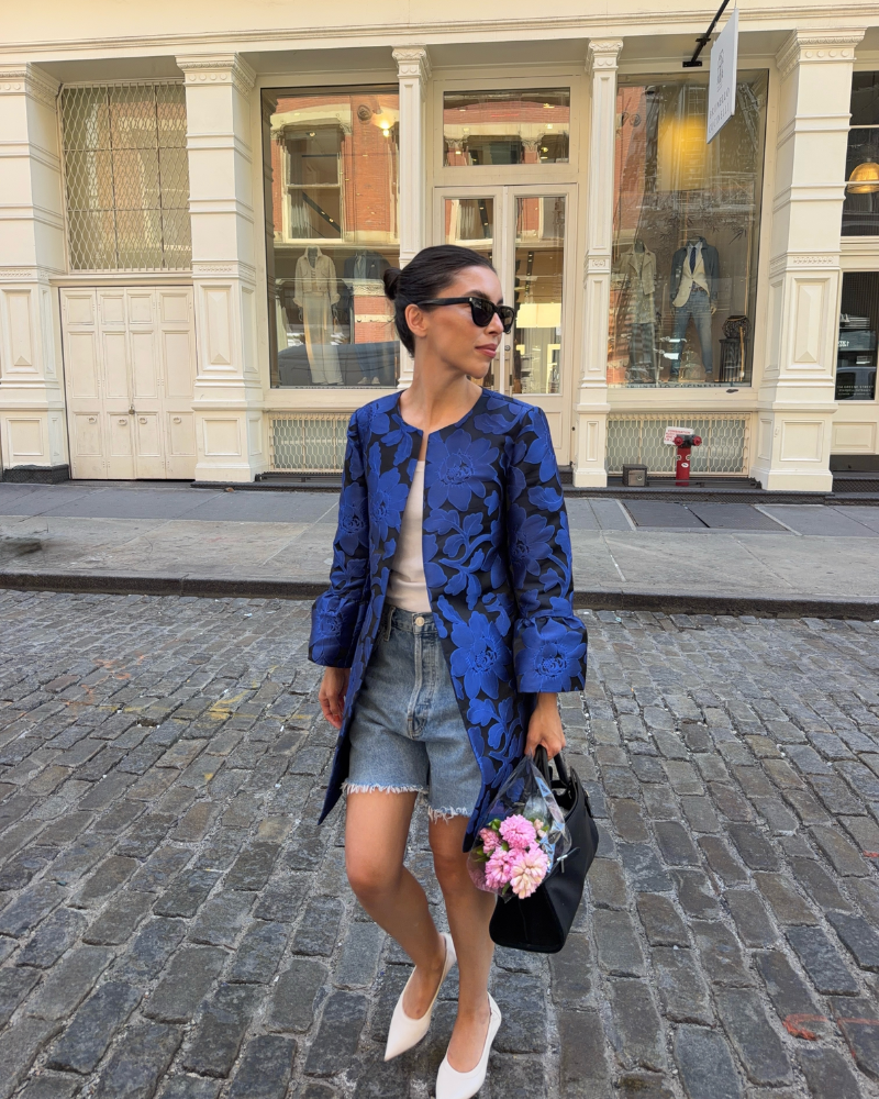 Chic outfit featuring a vibrant blue floral coat over a white top%2C paired with distressed denim shorts and sleek white heels%2C styled with a black handbag and a bouquet of pink flowers