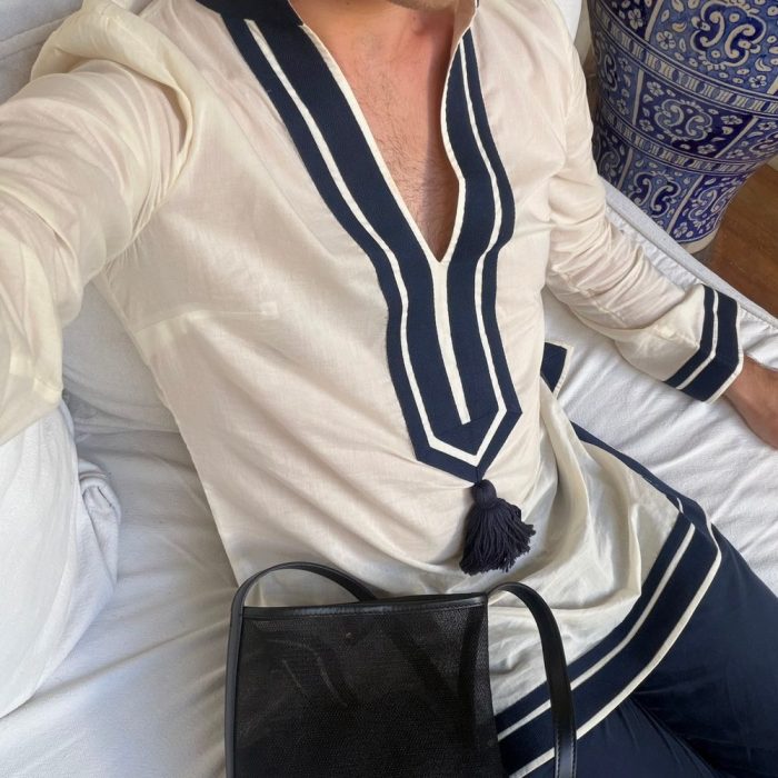 A person wearing a beige shirt with navy accents and tassel lounges on a white couch. A black bag rests nearby. Decorative blue vase in the background.