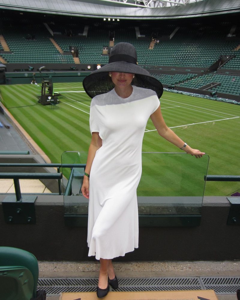 A chic white midi dress with a modern silhouette%2C paired with a wide-brimmed black hat and classic black flats%2C set against Wimbledon%27s iconic grass court backdrop