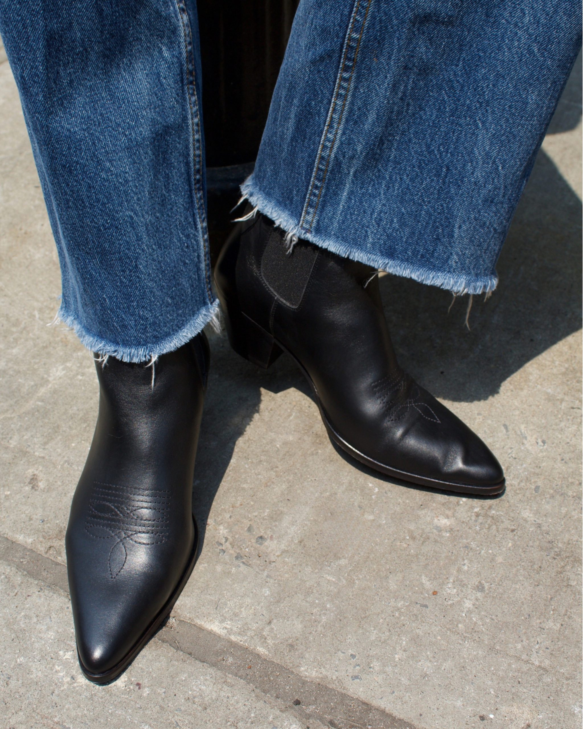 Black leather Nili Lotan ankle boots with pointed toes and subtle stitching paired with frayed hem blue denim jeans on concrete floor