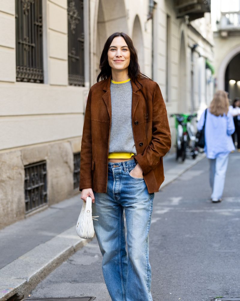 Woman wearing a brown suede blazer over a gray and yellow sweater with loose blue jeans and black ballet flats holding a white woven handbag on a city street