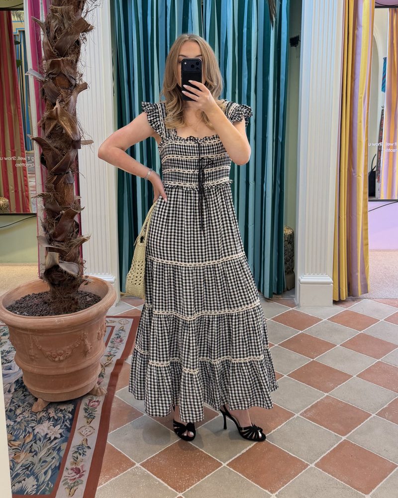 Rixo summer dress features a black and white gingham pattern with ruffled sleeves and tiered skirt%2C styled with heeled sandals and a woven bag.