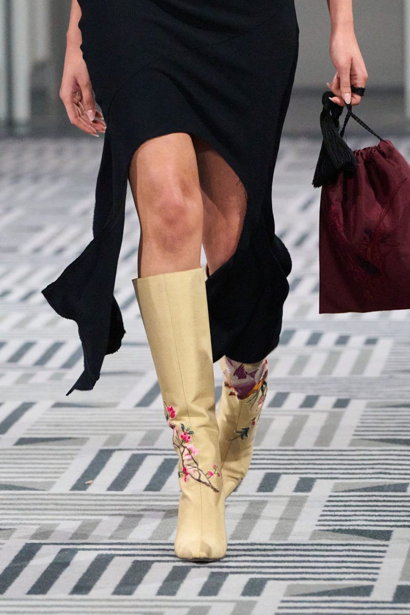Black asymmetrical slit dress paired with beige knee-high boots featuring floral embroidery and a burgundy tassel drawstring bag