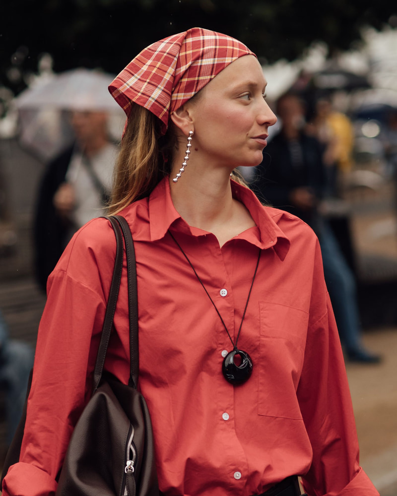 Woman wearing a red button-up shirt with rolled sleeves%2C plaid headscarf%2C black wide-leg trousers%2C black leather shoulder bag%2C and holding a transparent umbrella on a rainy street