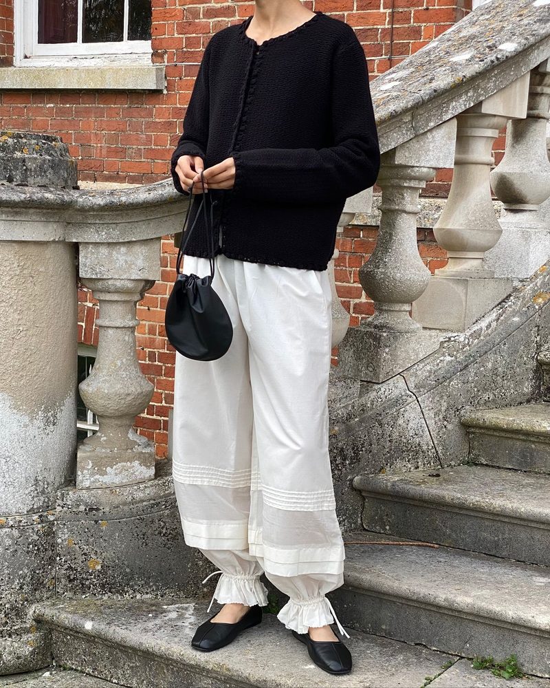 Black textured knit cardigan paired with loose white cotton trousers featuring sheer panel and ruffled cuffs%2C styled with black leather pouch bag and flats