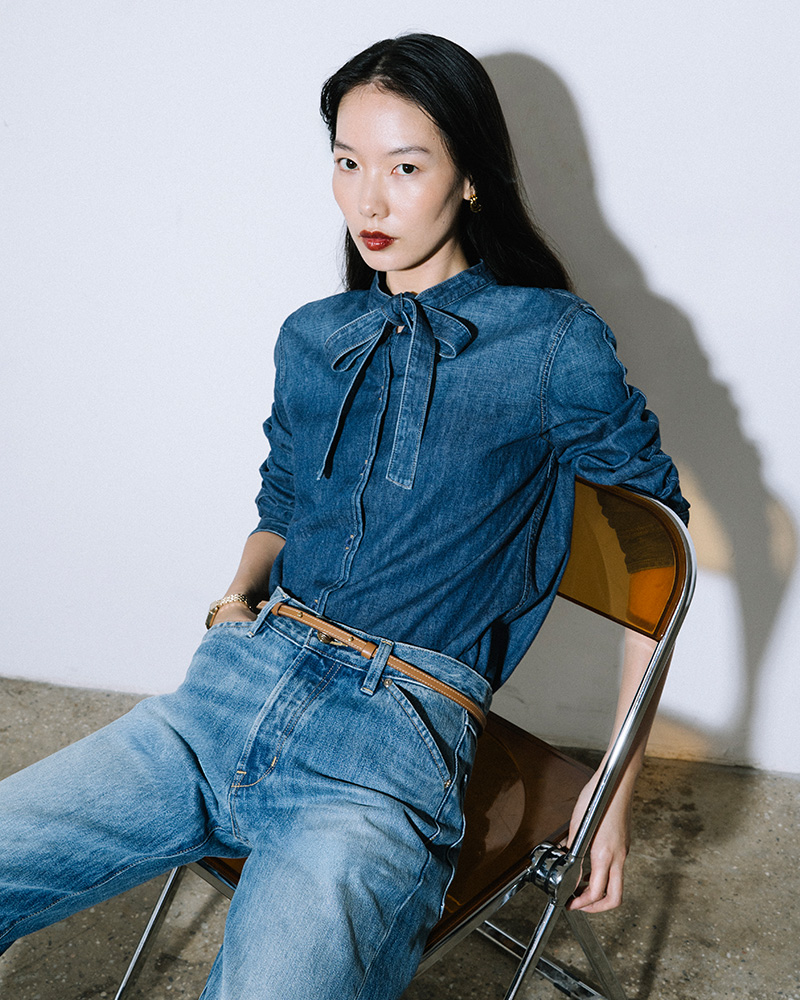 Denim bow-neck shirt styled with light-wash jeans and a tan belt%2C seated against a minimalist studio backdrop