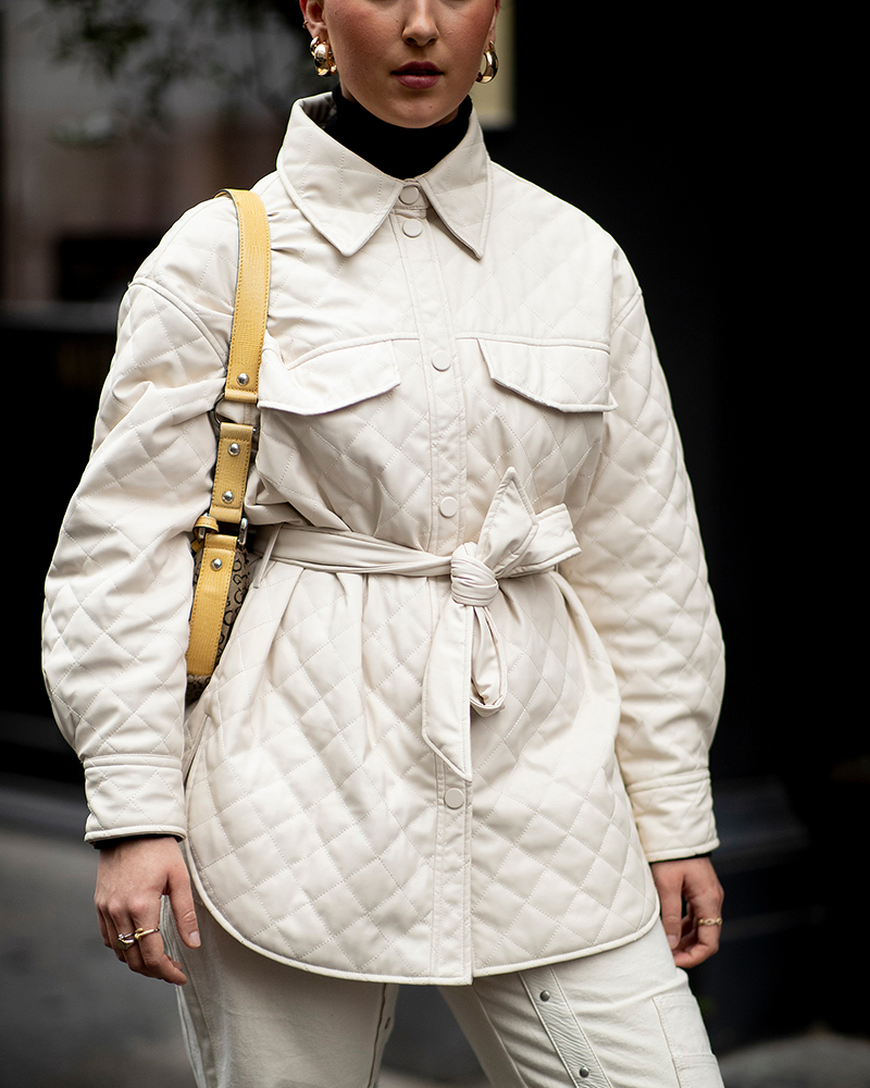 Model in an ivory quilted belted jacket with gold earrings and a yellow shoulder bag on a city street